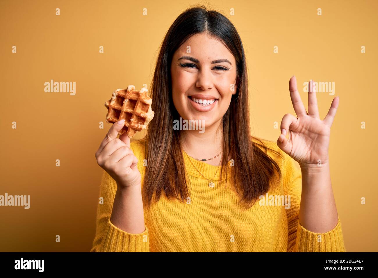 Young beautiful woman eating sweet waffle pastry over yellow background ...