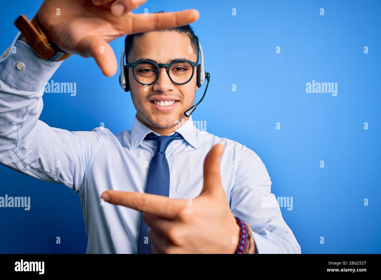 Young brazilian call center agent man wearing glasses and tie working using headset smiling ...