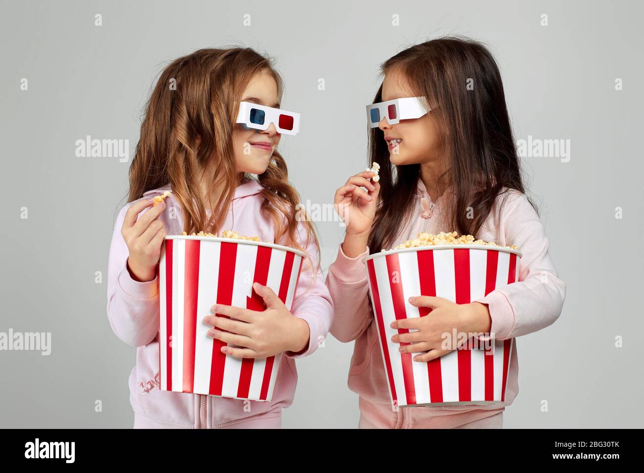 two beautiful caucasian little girls wearing red-blue 3d glasses and eating popcorn from bucket ...