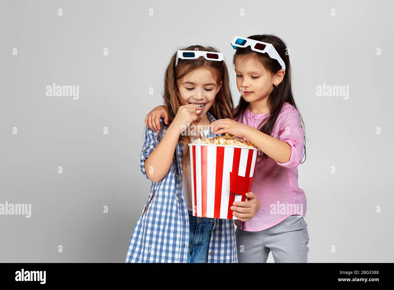 two beautiful caucasian little girls wearing red-blue 3d glasses and eating popcorn from bucket ...