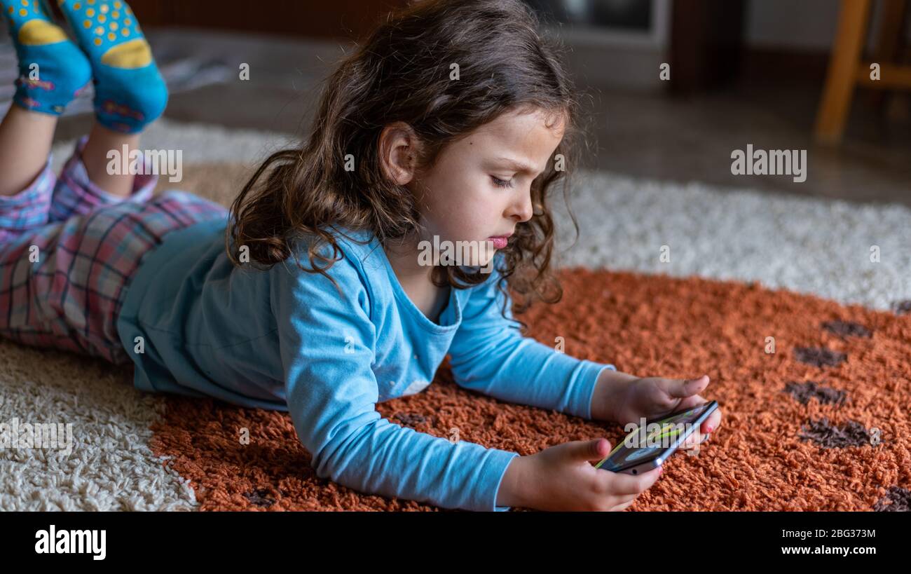Cute little happy girl lying down on using looking digital smartphone ...