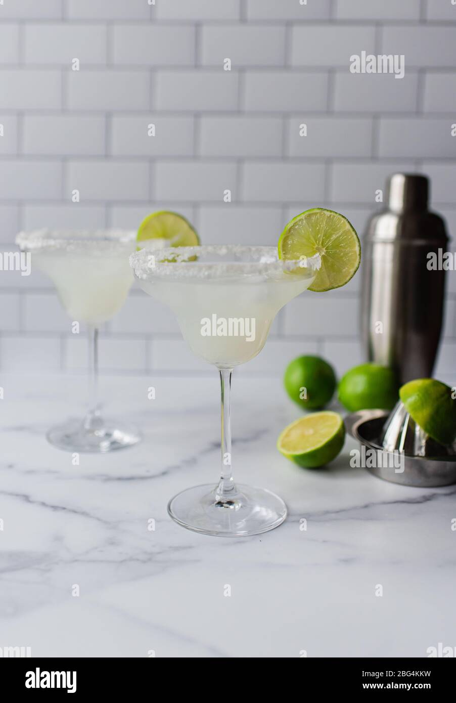Two margarita cocktails with limes and shaker on white marble counter ...