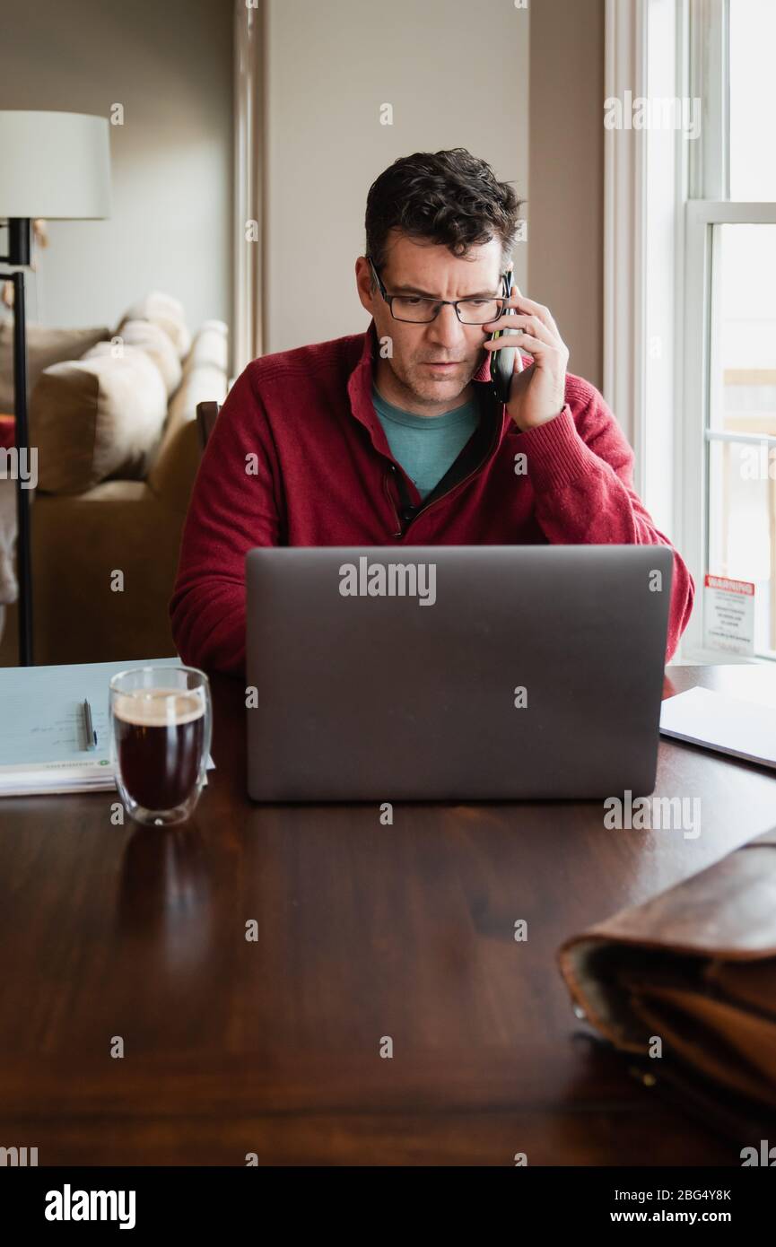 Man on cellphone working from home using a computer at a dining table ...