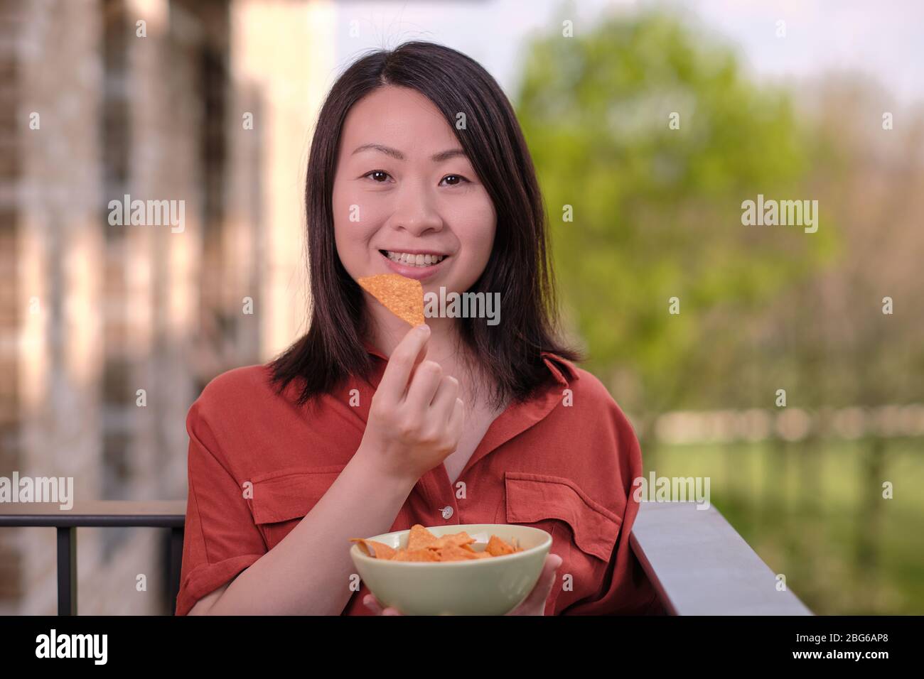Beautiful asian woman standing on balcony smiling and eating doritos ...