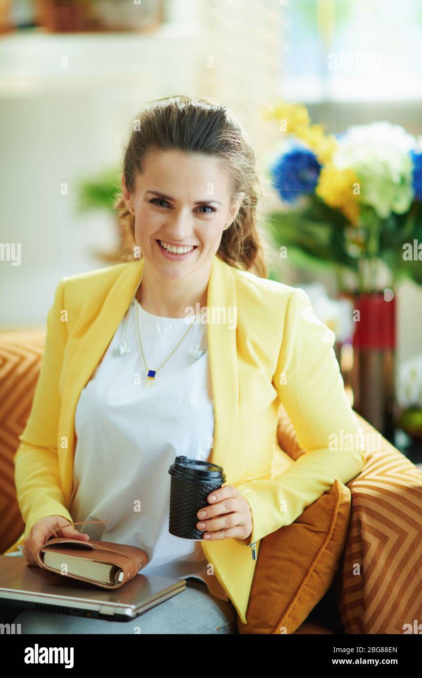 smiling trendy student woman in jeans and yellow jacket sitting on sofa ...