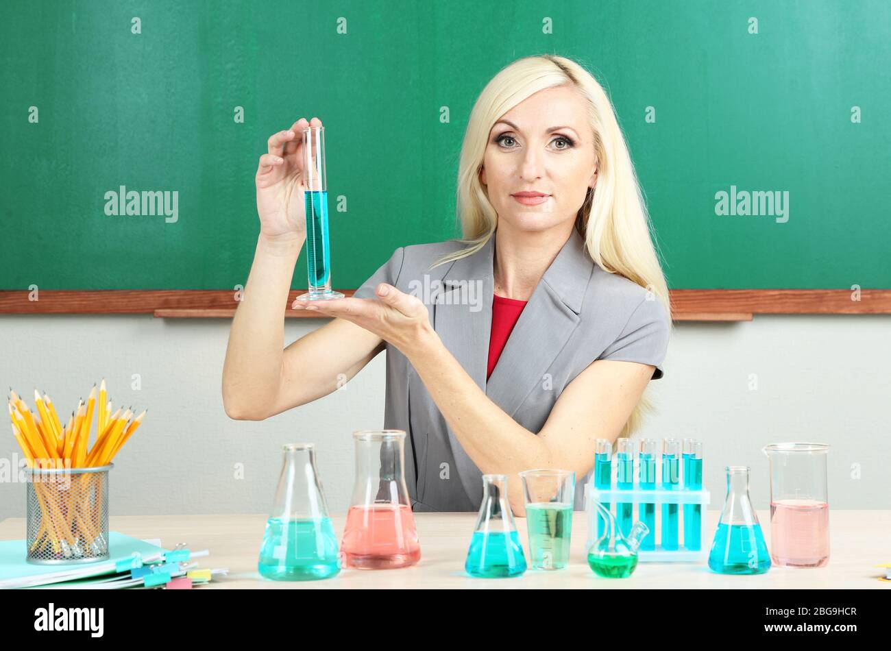 Chemistry teacher with tubes sitting at table on blackboard background ...