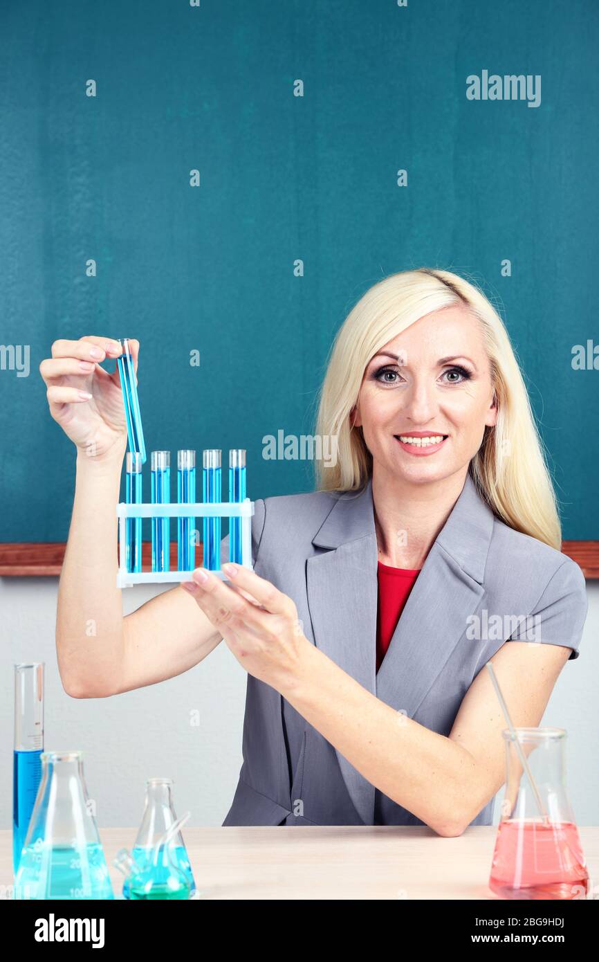 Chemistry teacher with tubes sitting at table on blackboard background ...