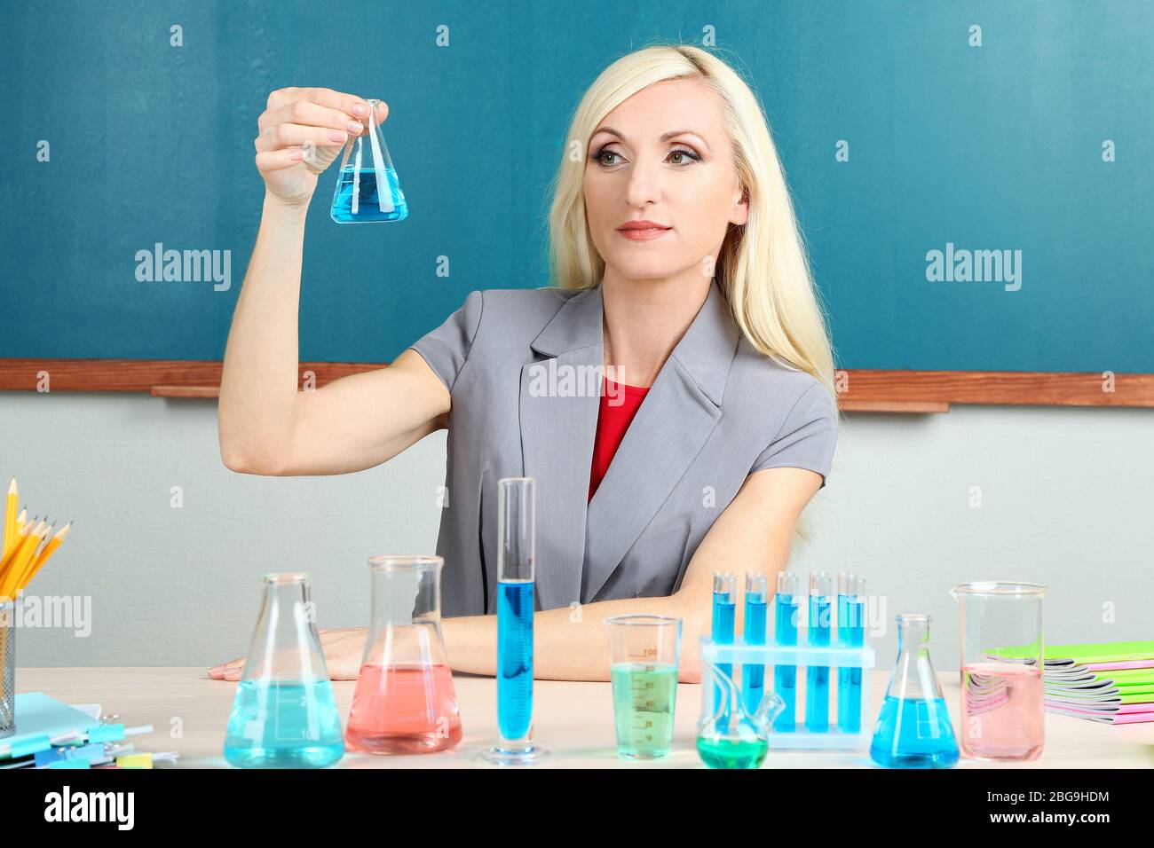 Chemistry teacher with tubes sitting at table on blackboard background ...