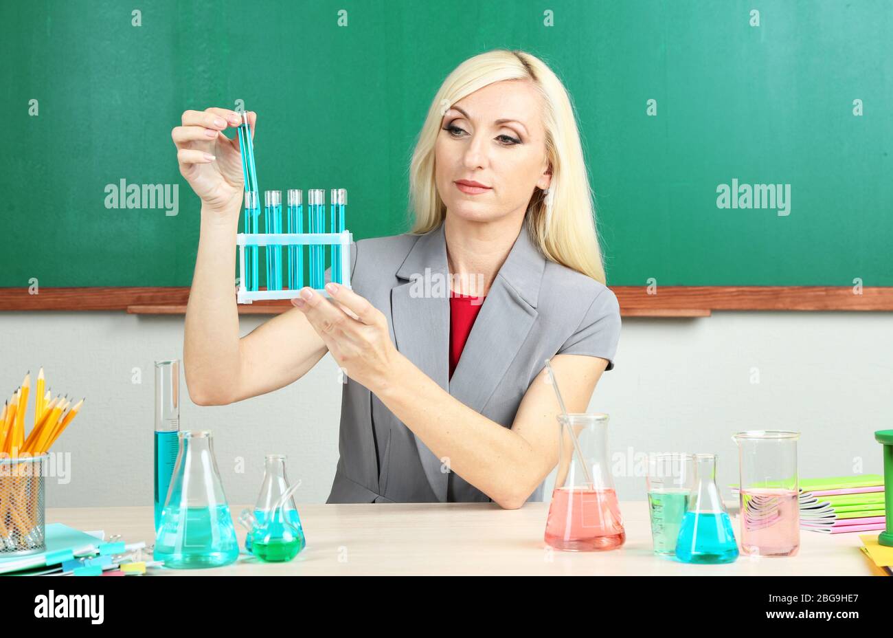 Chemistry teacher with tubes sitting at table on blackboard background ...