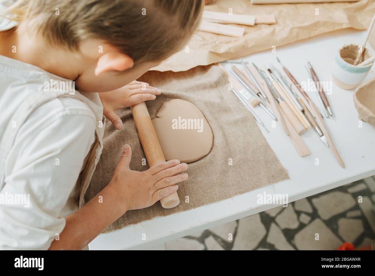 Boy rolling clay piece on a table filled with pottery tools Stock Photo ...