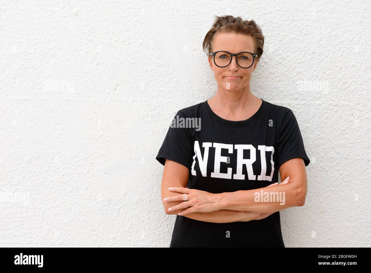 Serious mature nerd woman with arms crossed wearing big eyeglasses and standing against white ...