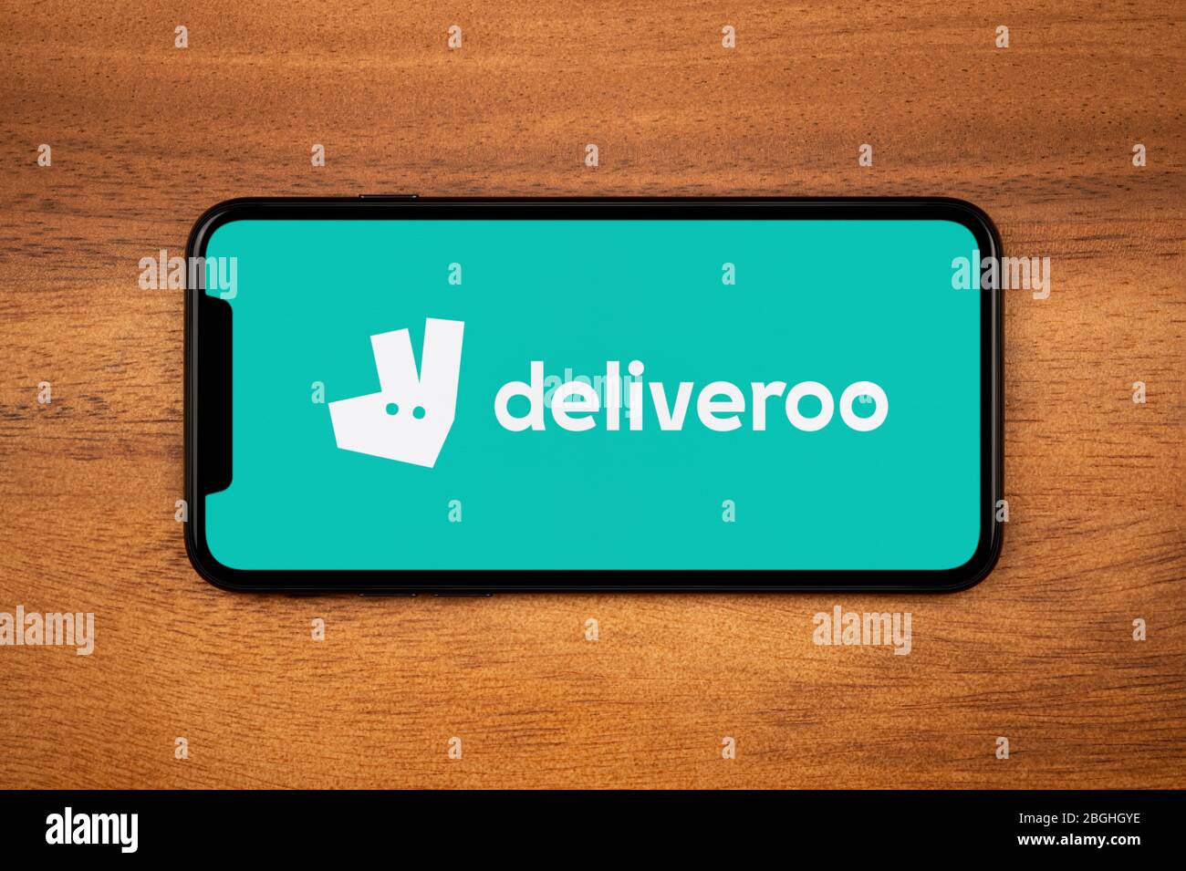 A smartphone showing the Deliveroo logo rests on a plain wooden table ...