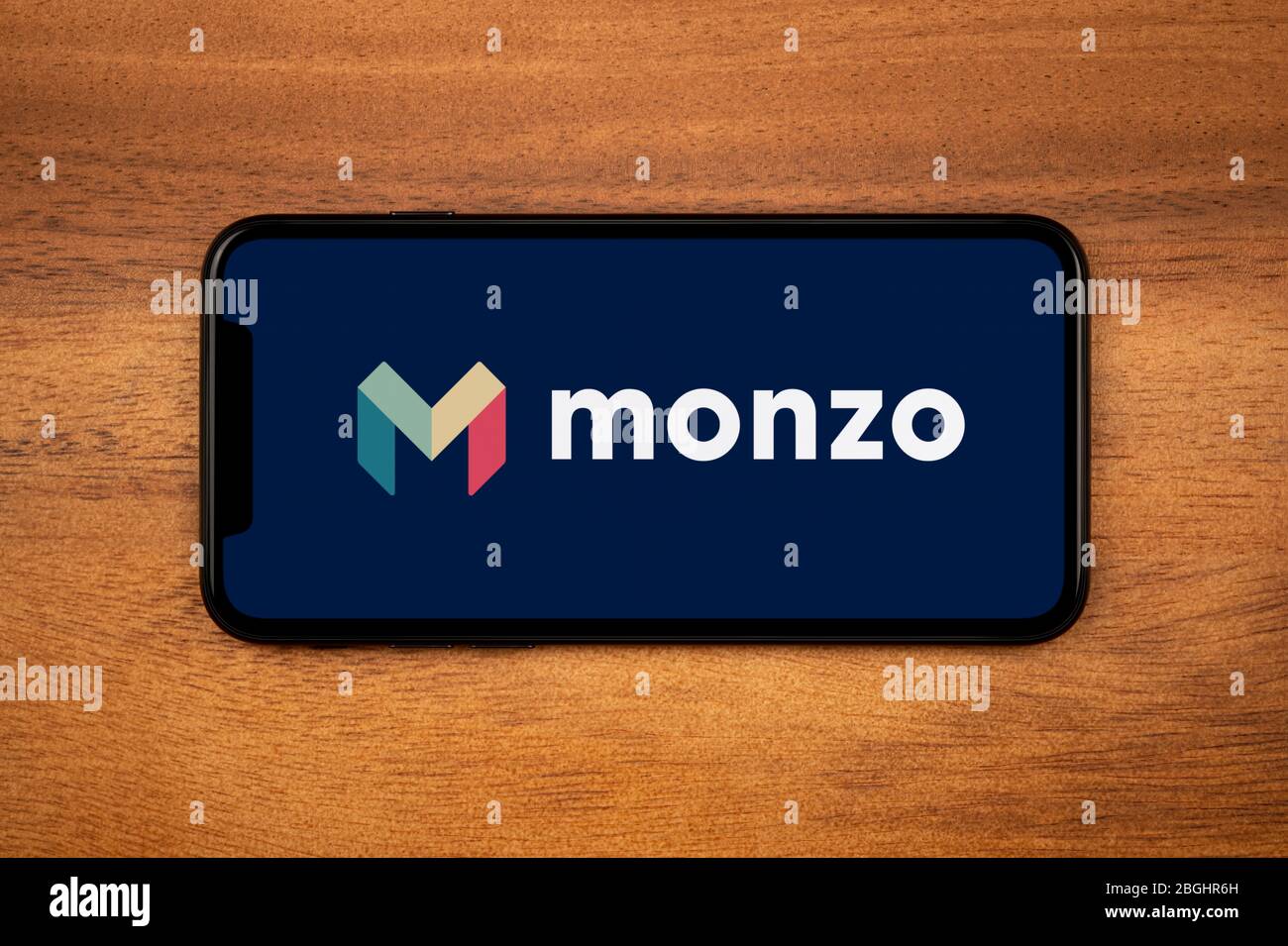 A smartphone showing the Monzo Bank logo rests on a plain wooden table ...