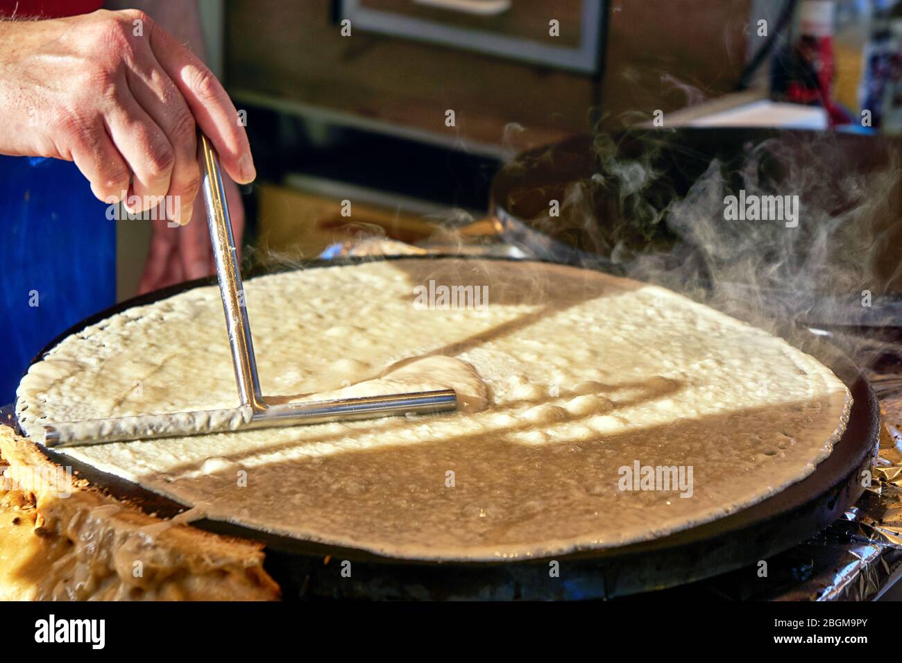 A hand makes crepes and pancakes on a metal grill with a metal stick ...