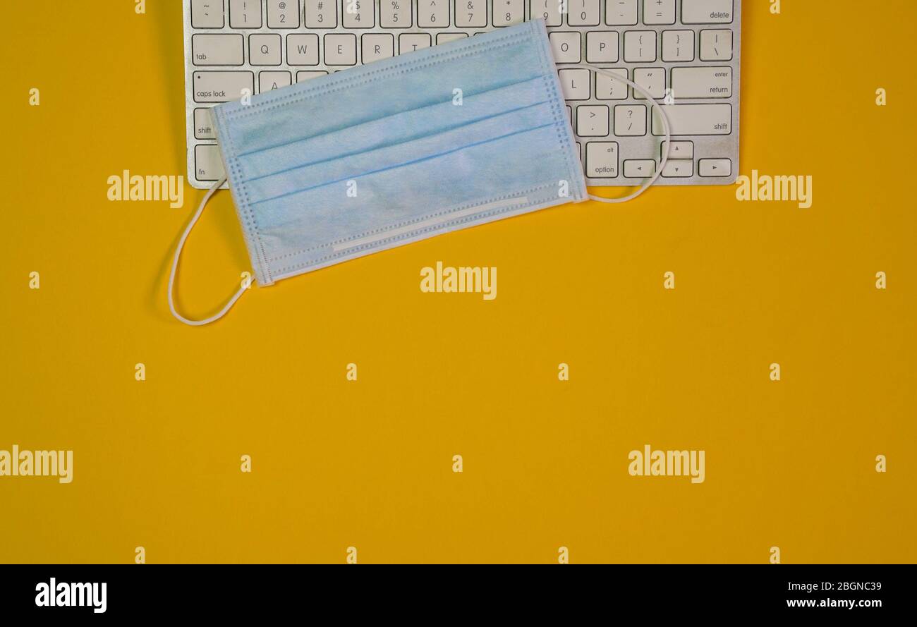 Medical face mask on top of keyboard. Yellow background. Top view. Copy ...