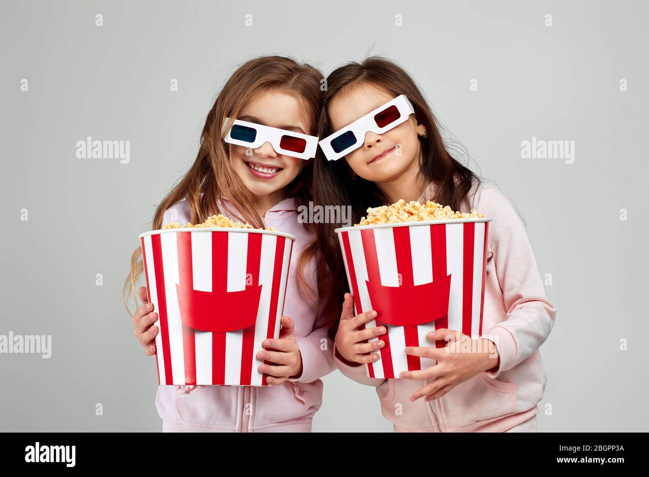 two beautiful caucasian smiling little girls in red-blue 3d glasses holding popcorn buckets and ...