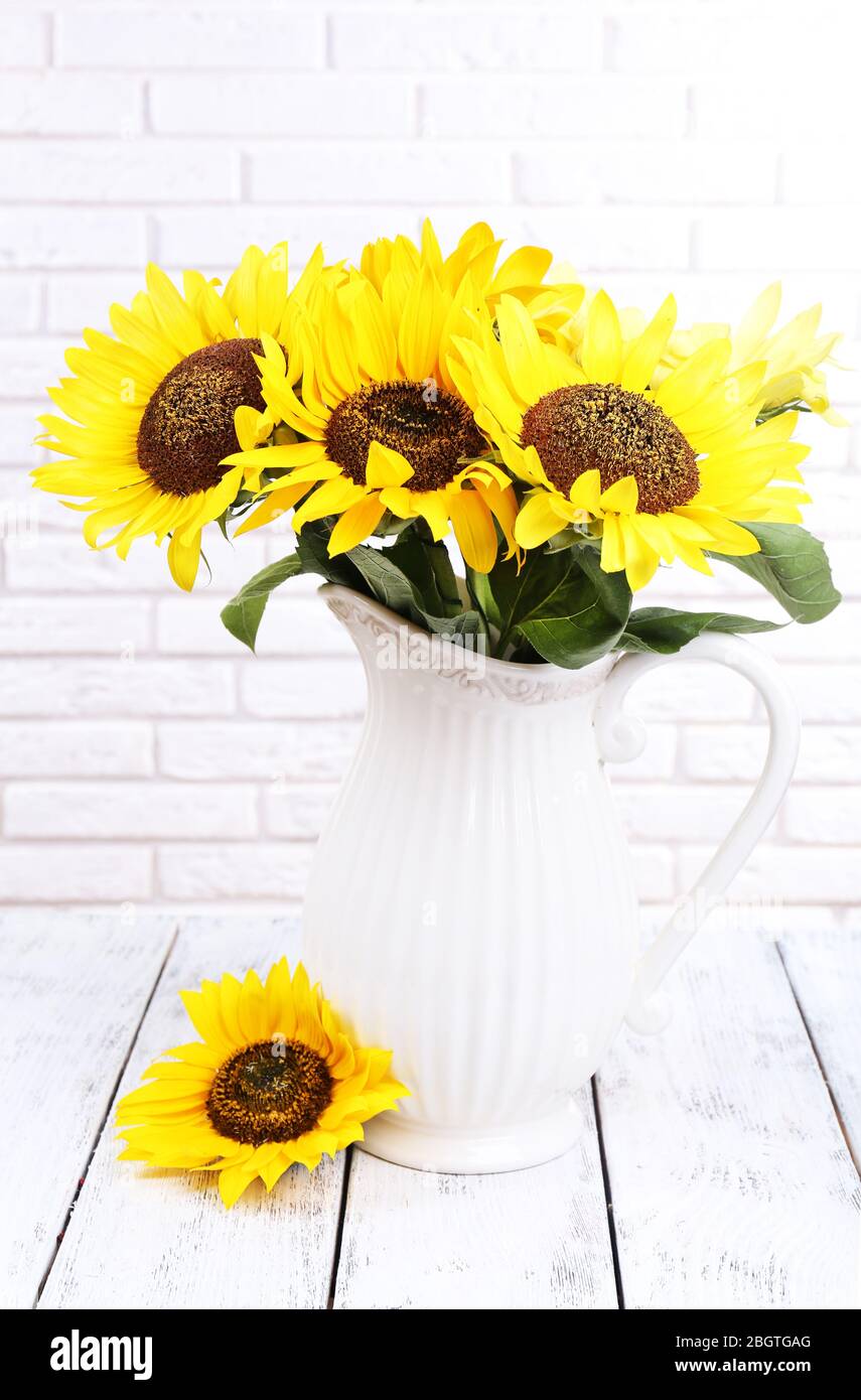 Beautiful bouquet of sunflowers in pitcher on table on brick wall ...