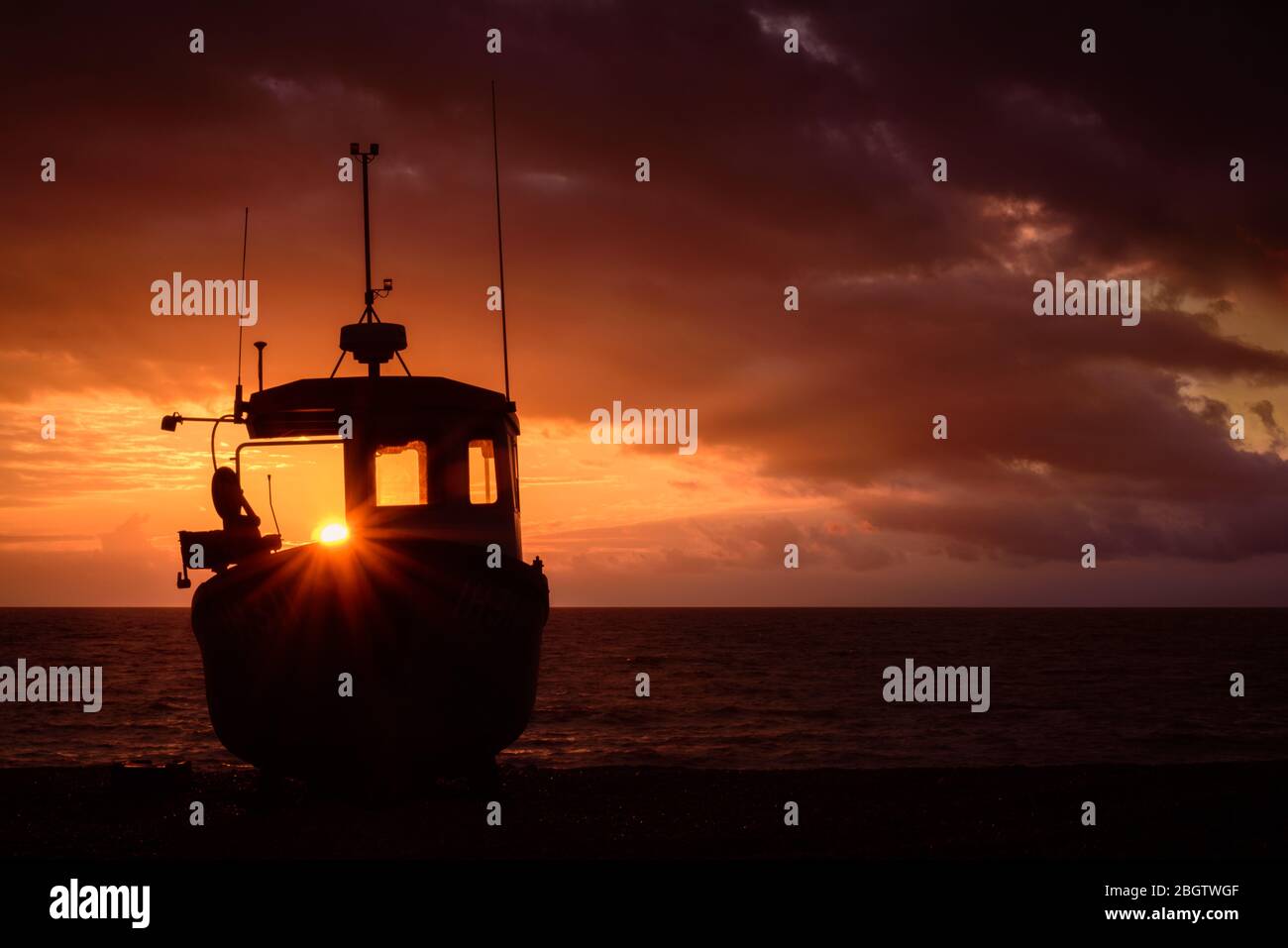 Fishing boat silhouette and sunburst on shingle beach at dawn with ...