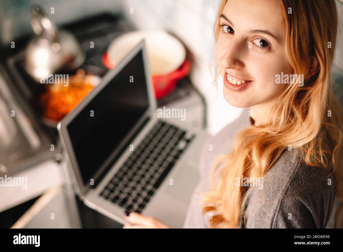 Happy young woman holding laptop computer while cooking food on kitchen ...