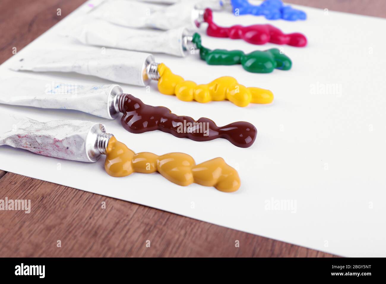 Colorful paint tubes squeezed on white sheet of paper and wooden table ...