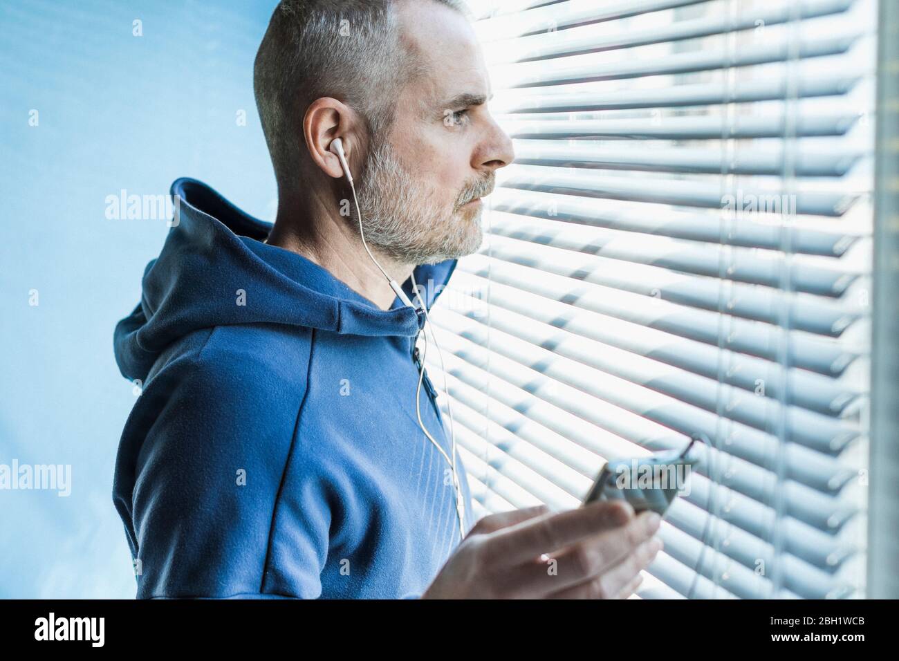 Pensive man with smartphone and earbuds looking out of venetian blind ...