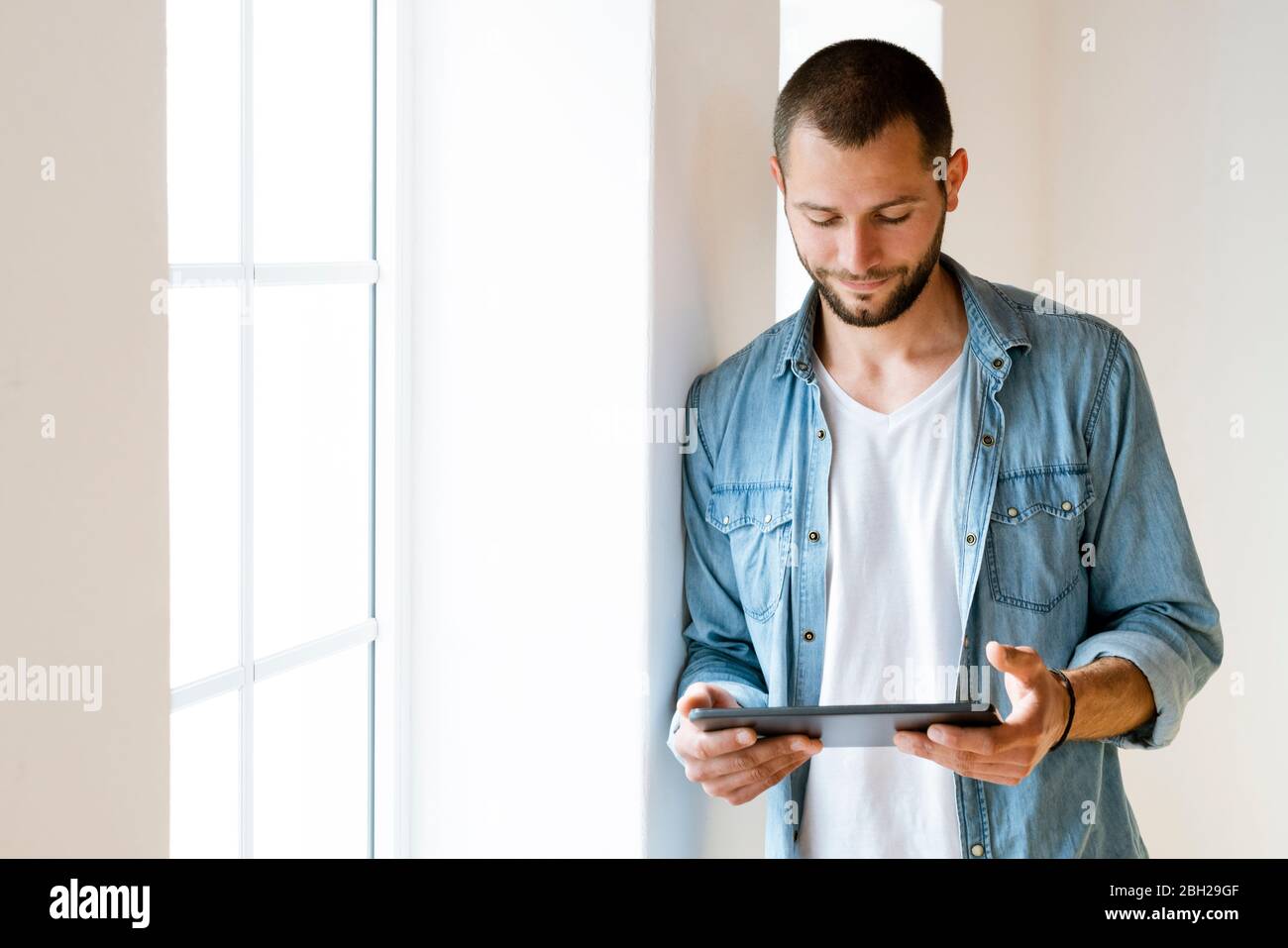 Young man at home smiling and looking at his tablet standing at window ...