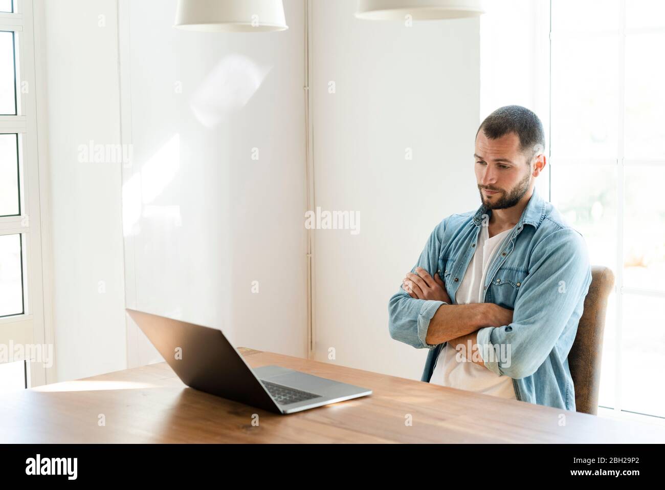 Serious young man with crossed arms at home looking at his laptop in ...
