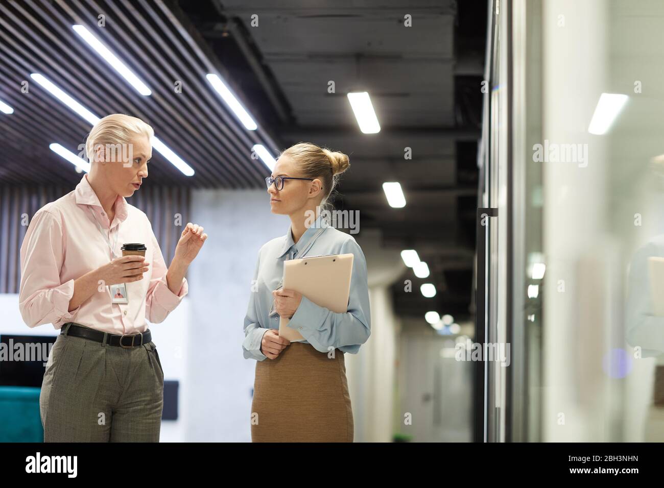 Two colleagues talking to each other while standing at office corridor ...