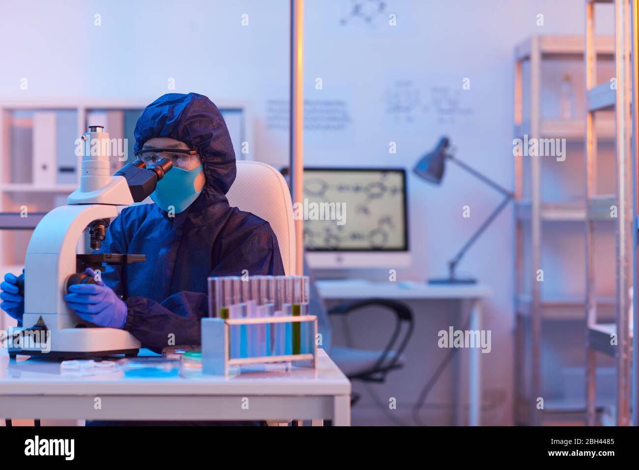 Scientist in protective workwear and mask sitting at her workplace with ...