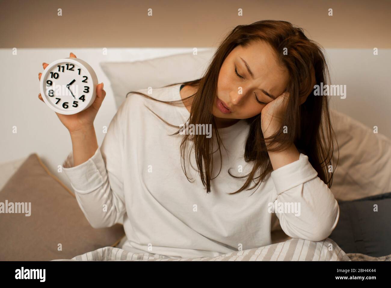 Tired pretty girl taking a nap sitting in bed with alarm clock in her ...