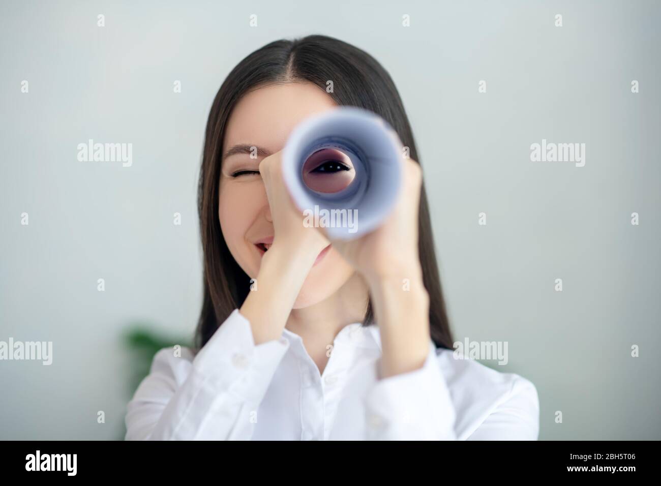 At work. Young brunette female looking through paper tube, smiling ...
