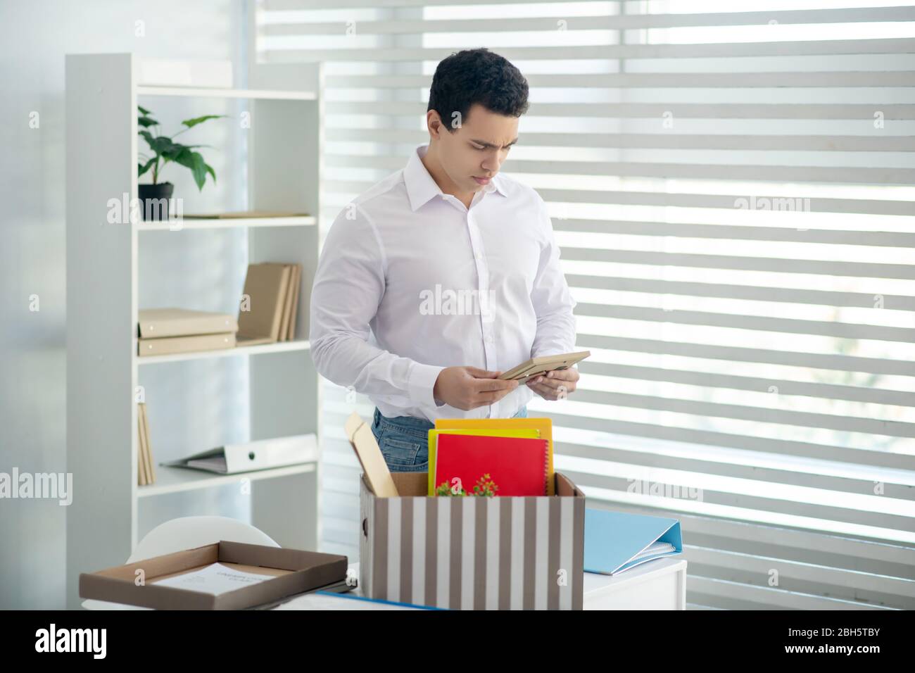 At work. Male standing at his desk with box of personal stuff, holding ...