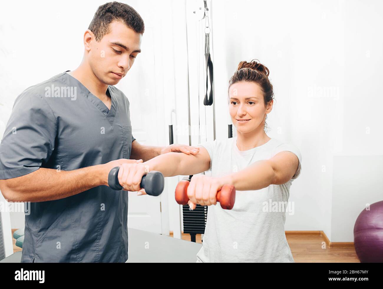 Physiotherapist working with male patient in clinic. Raises dumbbells ...