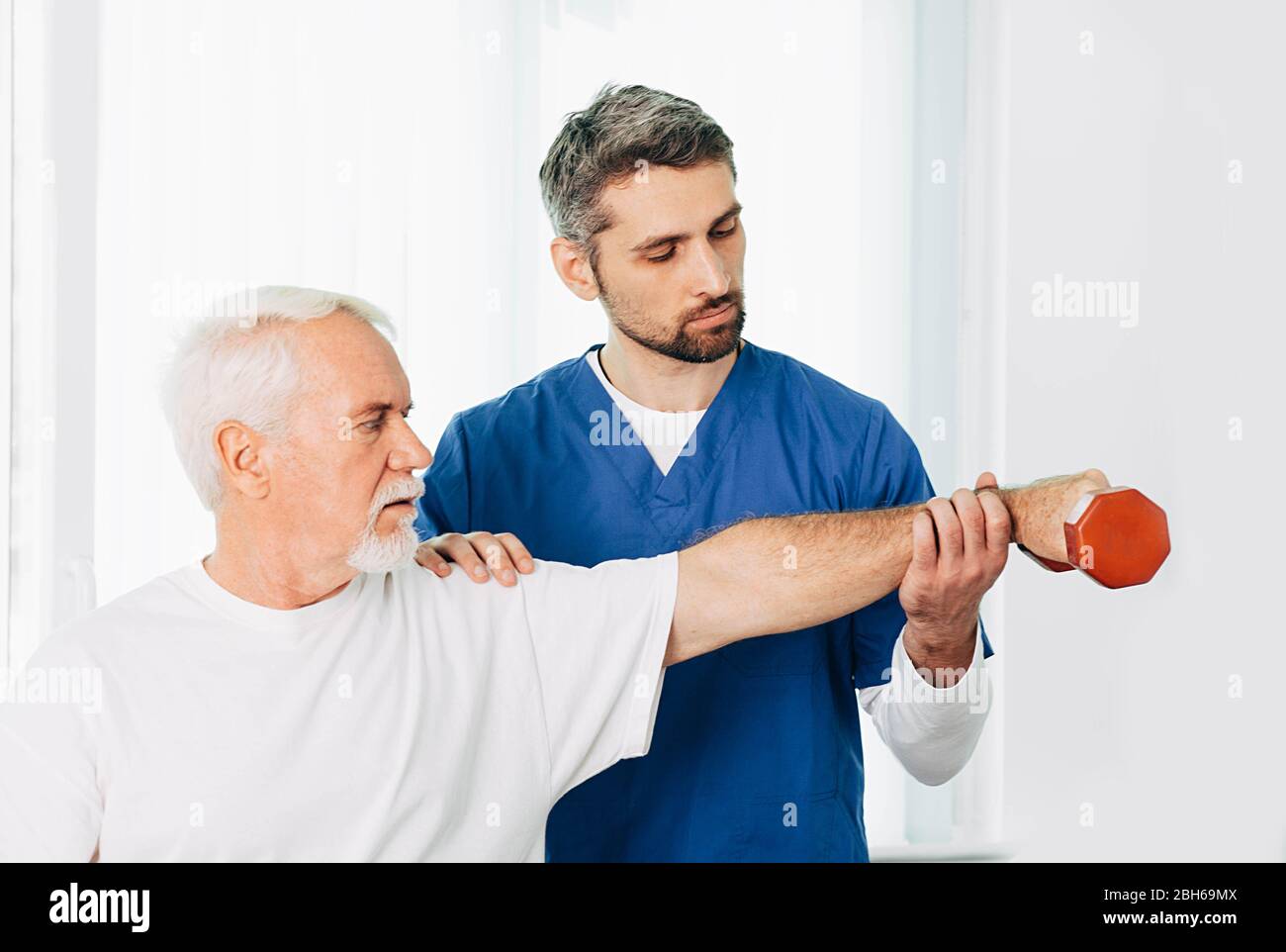 Positive physiotherapist helping his senior patient to lift a dumbbell ...