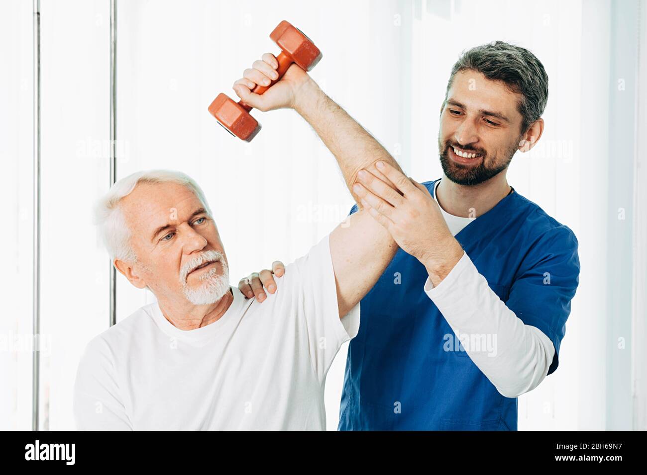 Positive physiotherapist helping his senior patient to lift a dumbbell ...