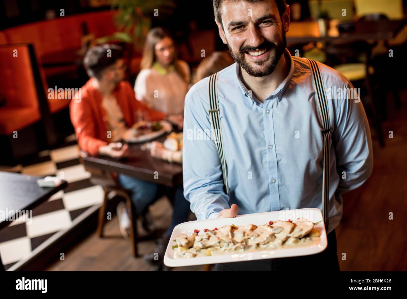 Smiling young waiter carrying plate with meat dish in the restaurant ...