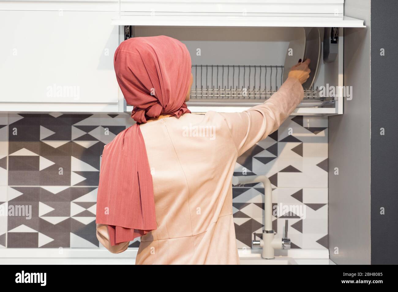 muslim woman in hijab opens kitchen cabinet on modern kitchen back view ...