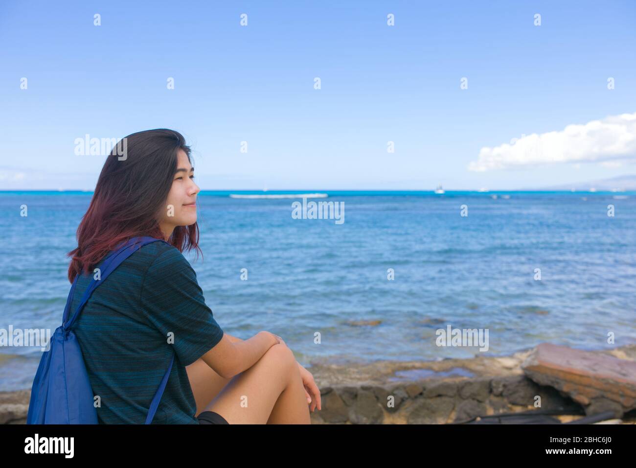 Smiling biracial Asian teen girl sitting alone along Hawaiian ocean ...