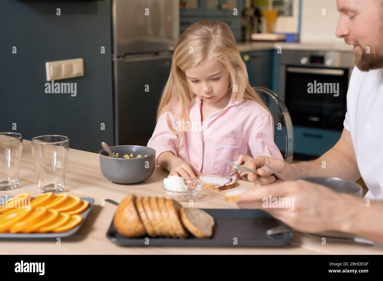 Pretty blond little girl spreading butter on slice of wheat bread while ...