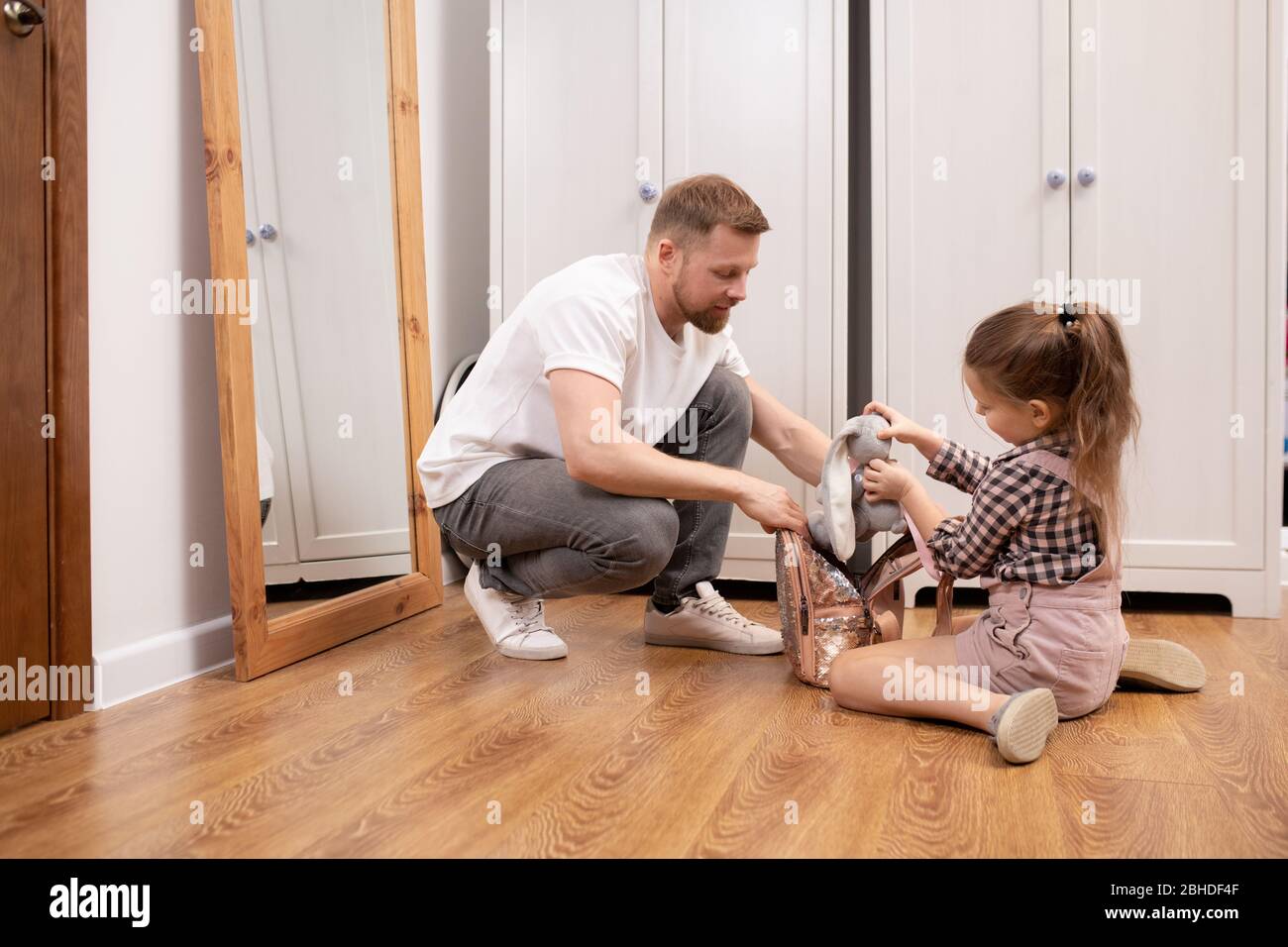 Cute little girl putting soft toy into rucksack while father helping ...