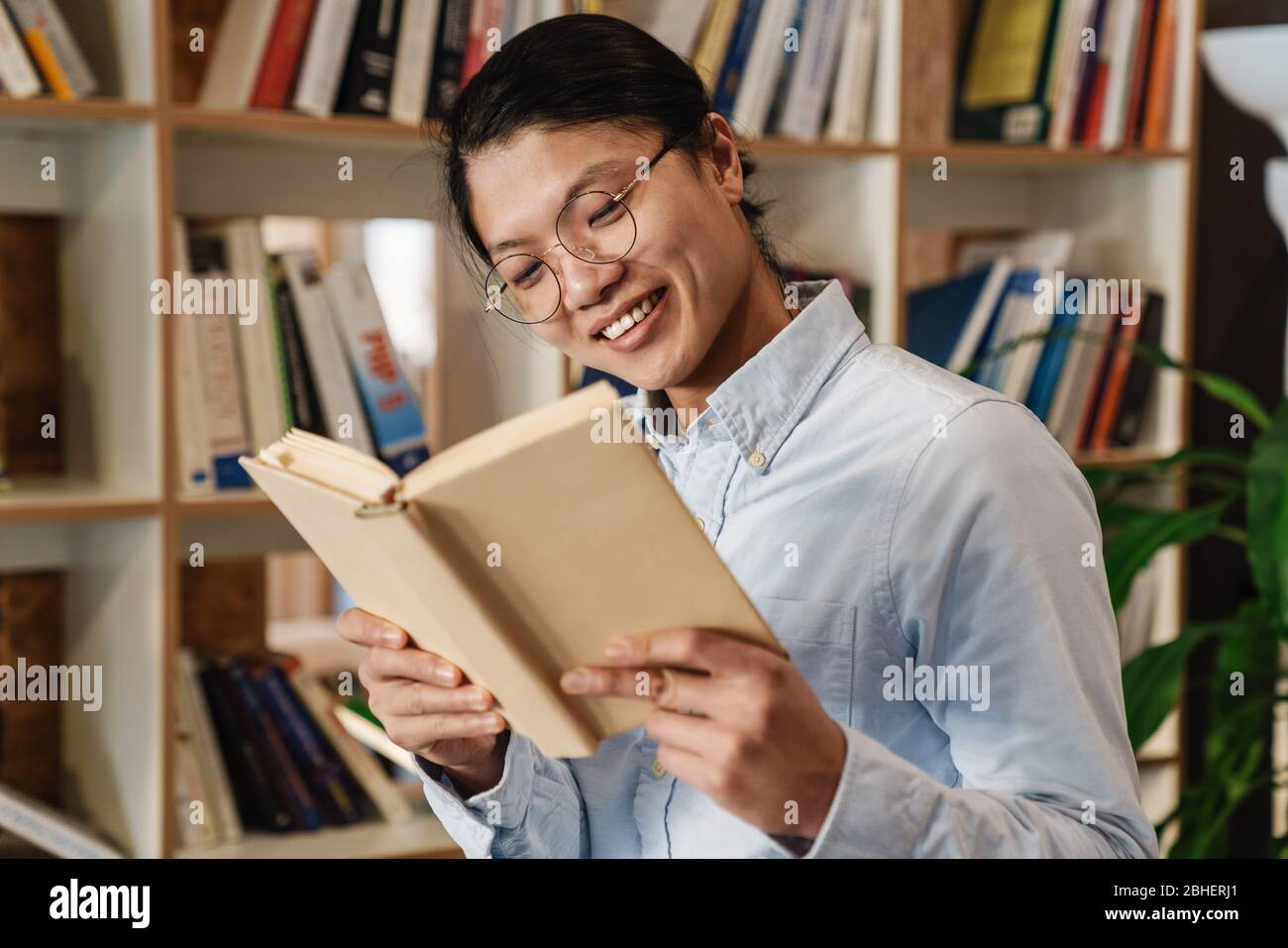 Image of joyful handsome asian man in eyeglasses smiling and reading ...