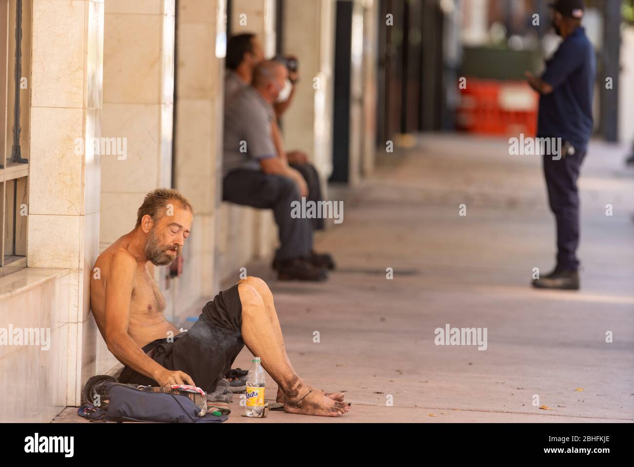 Homeless man sitting on the streets among wokring men in the background ...