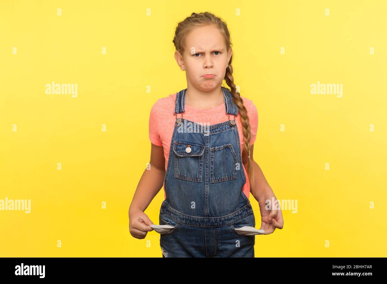 Portrait of upset little girl with braid in denim overalls turning out ...