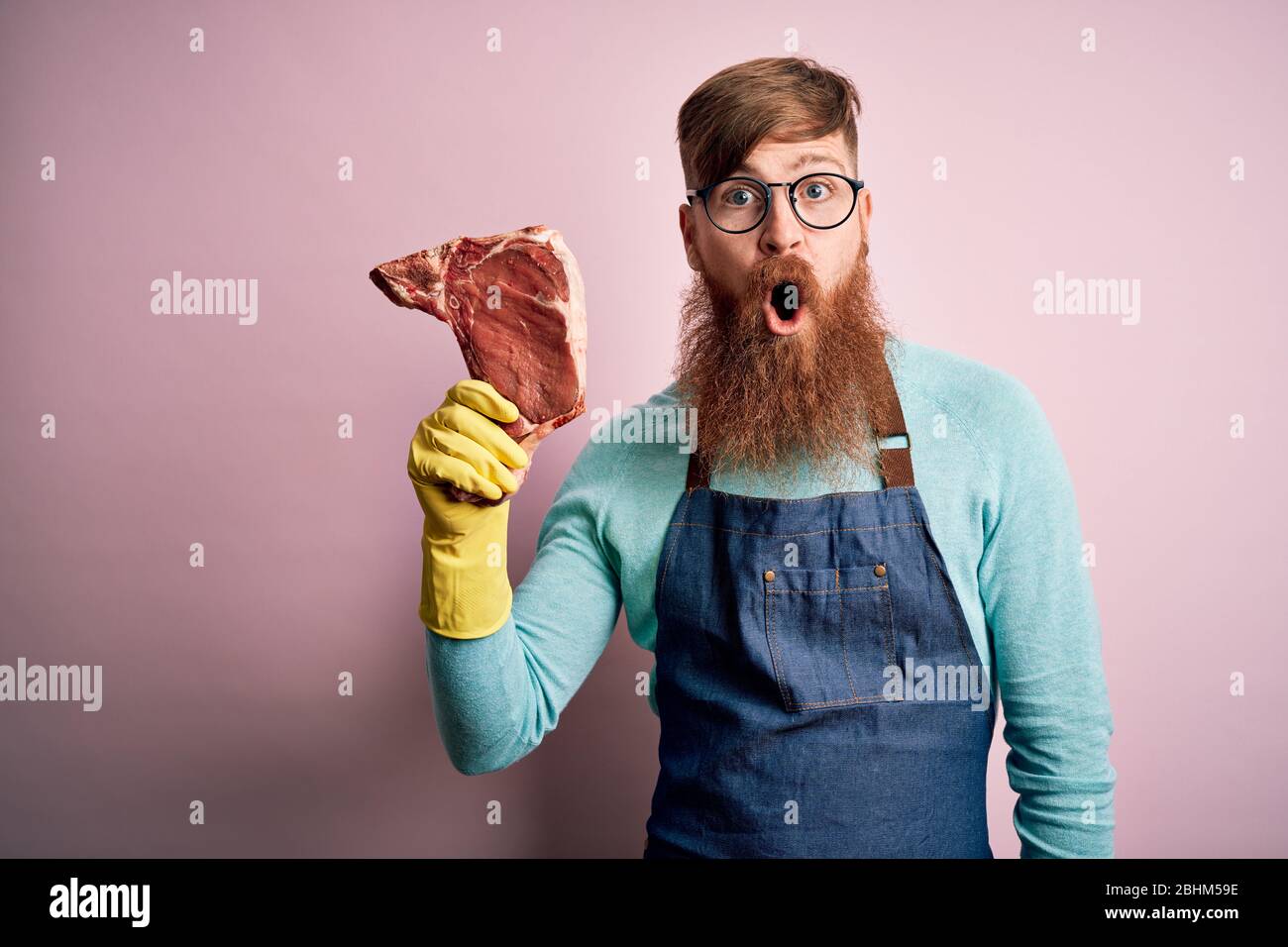 Redhead Irish butcher man with beard holding raw beef steak over pink ...