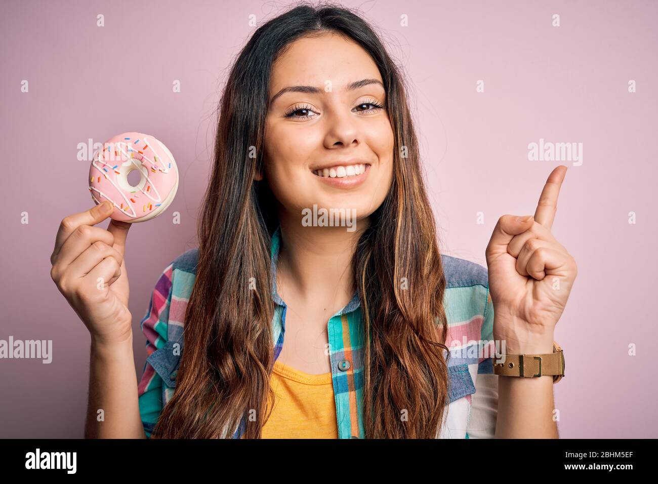 Young beautiful brunette woman eating sweet pink doughnut over isolated ...