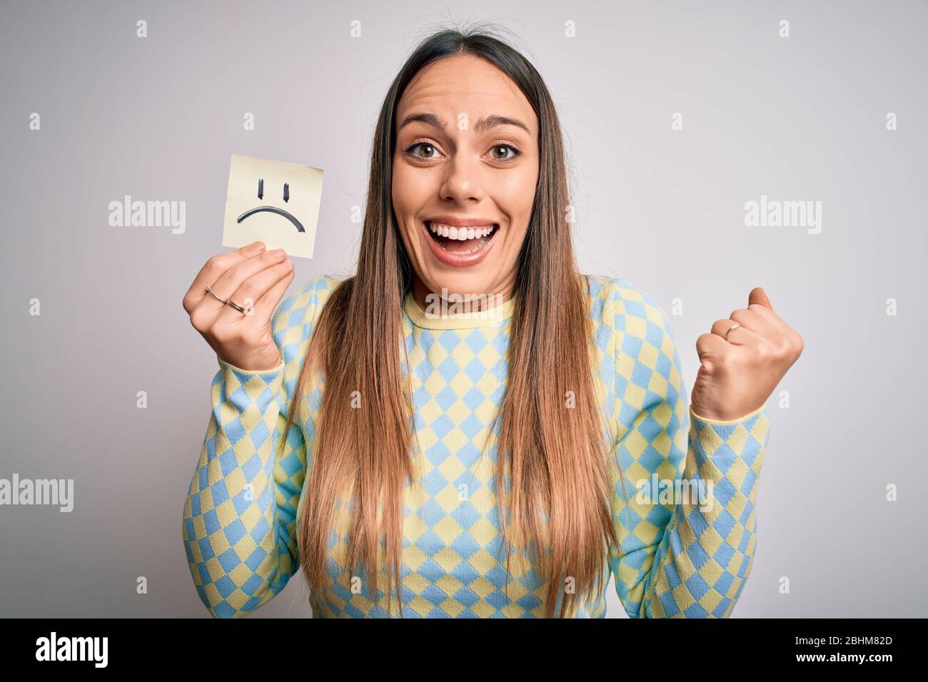 Young blonde woman holding sticky paper note with sad face emoticon ...
