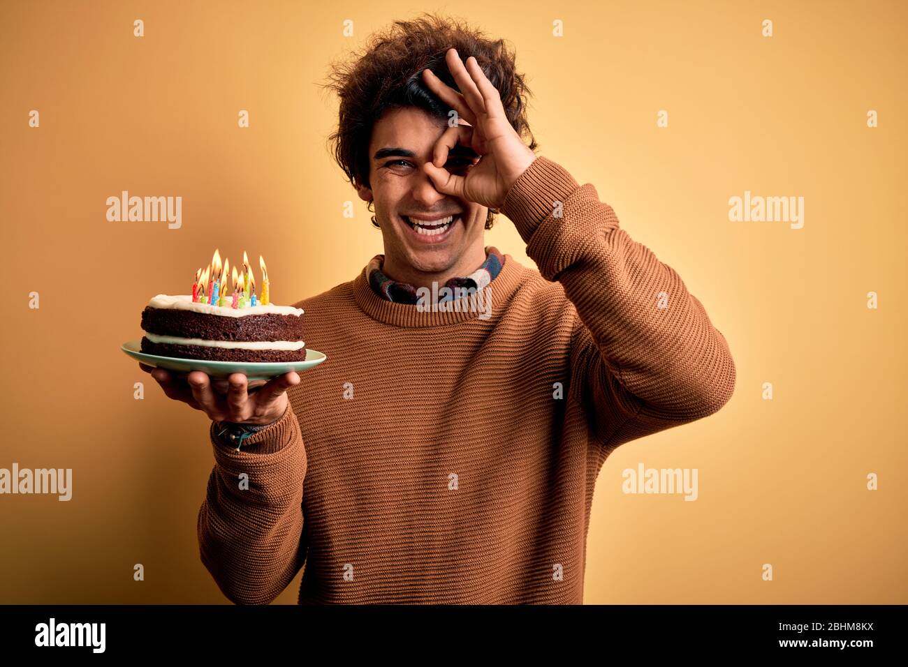 Young handsome man holding birthday cake standing over isolated yellow ...