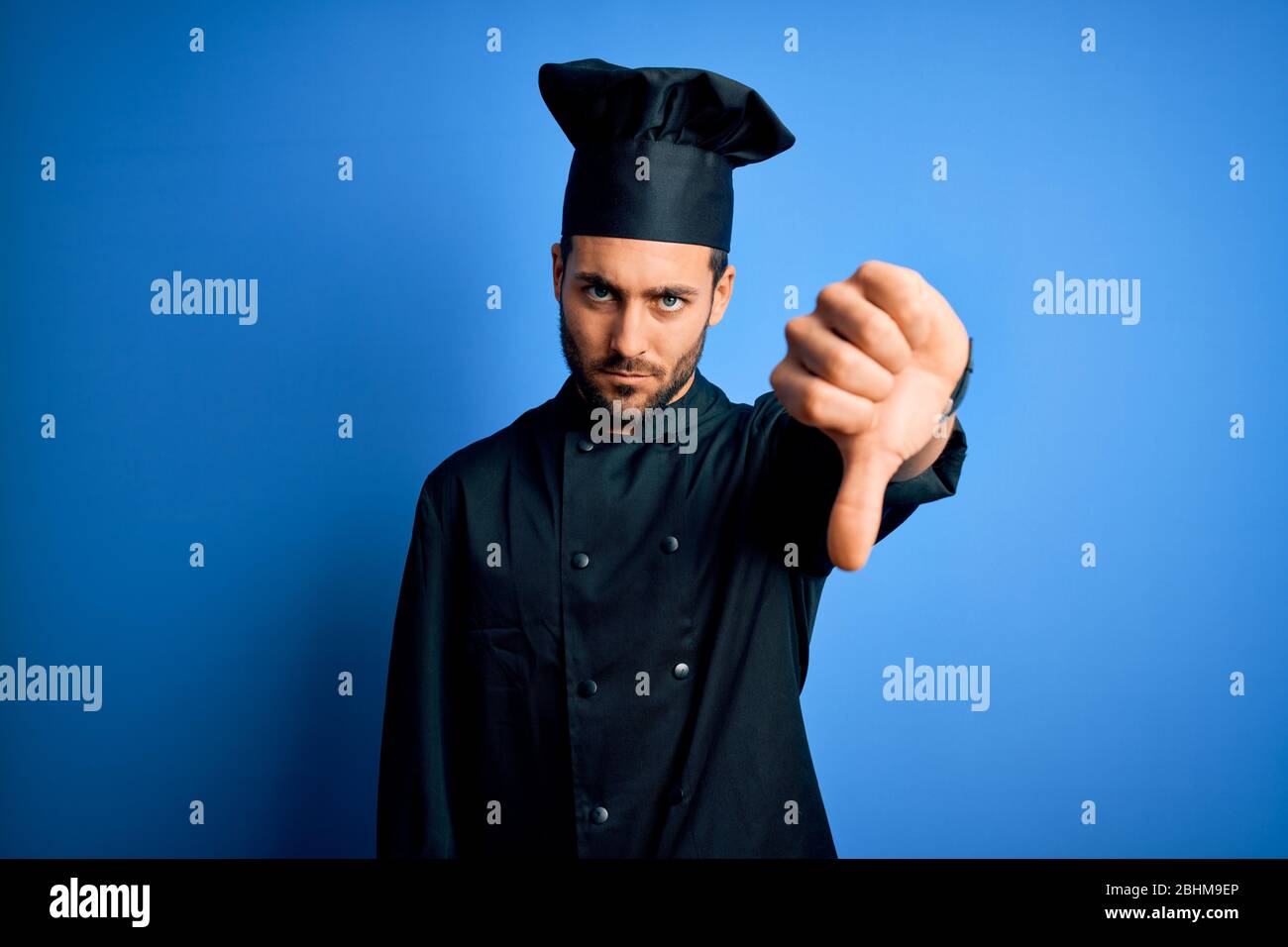 Young handsome chef man with beard wearing cooker uniform and hat over ...