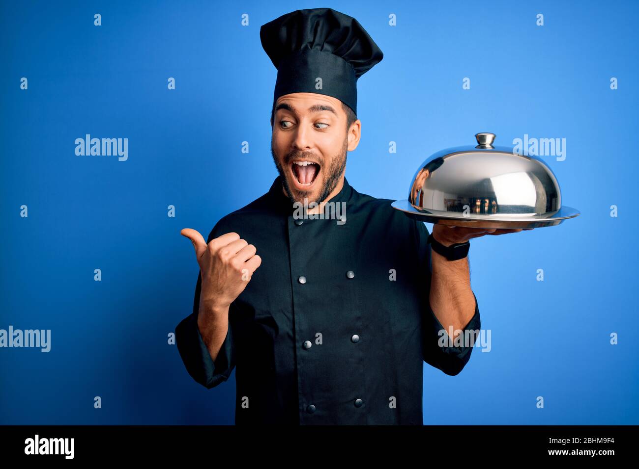 Young cooker man with beard wearing uniform holding tray with dome over ...
