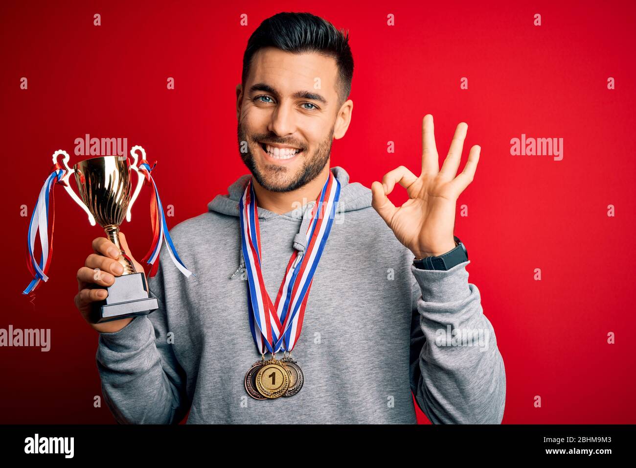 Young handsome succesful man holding trophy wearing medals over red ...